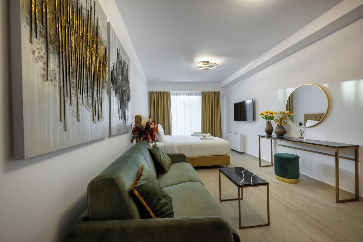 Quarto de hotel moderno com sofá verde, uma cama, televisão, mesa consola e um grande quadro abstrato.