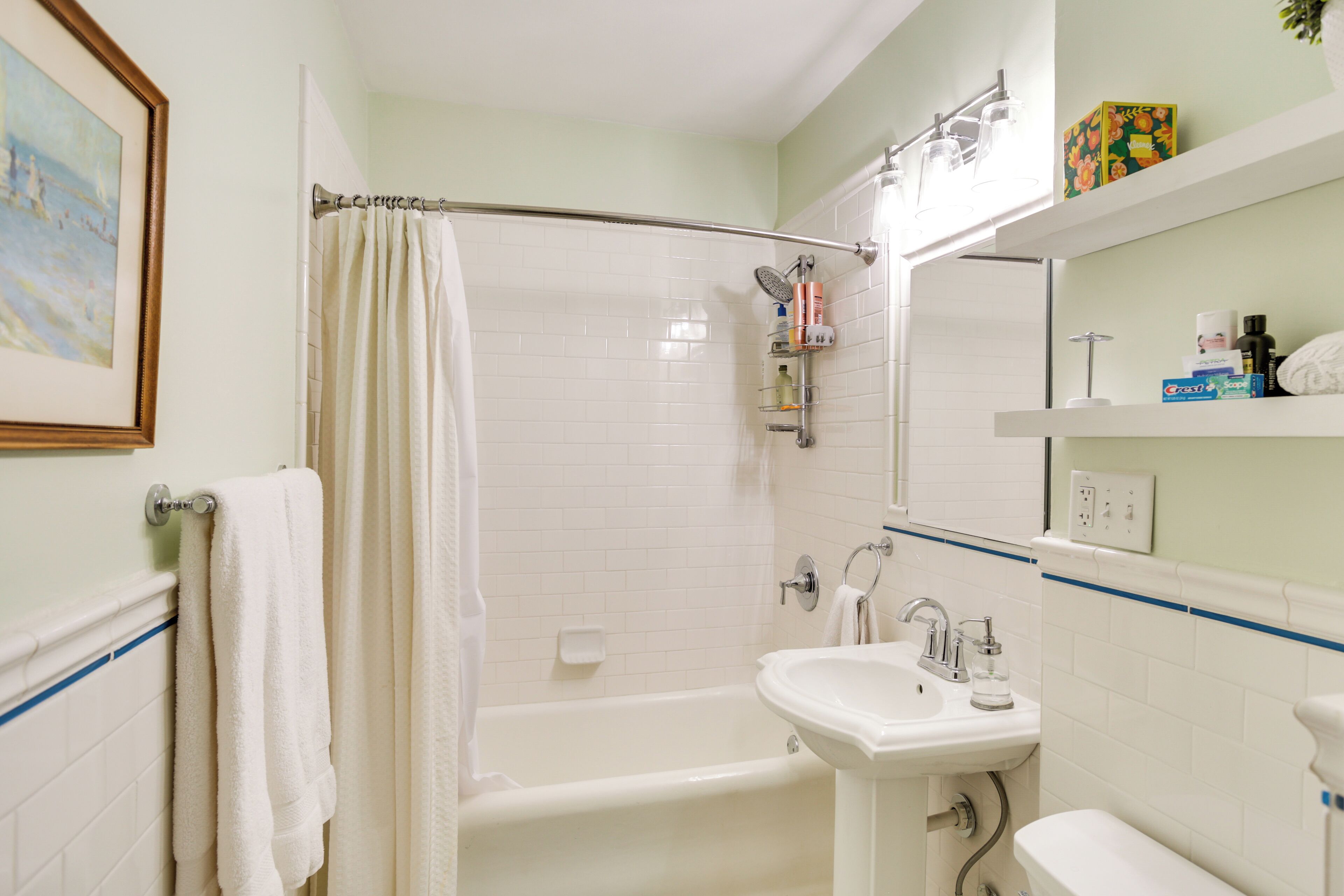 Uma casa de banho com banheira, lavatório e sanita. Existem prateleiras com produtos de higiene pessoal e um quadro emoldurado na parede.