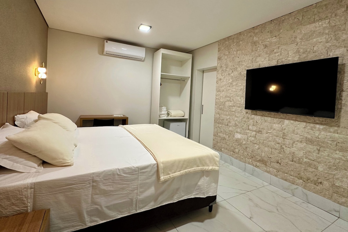 Quarto de hotel com cama, televisão na parede, ar condicionado, mesa de cabeceira e guarda-roupa.