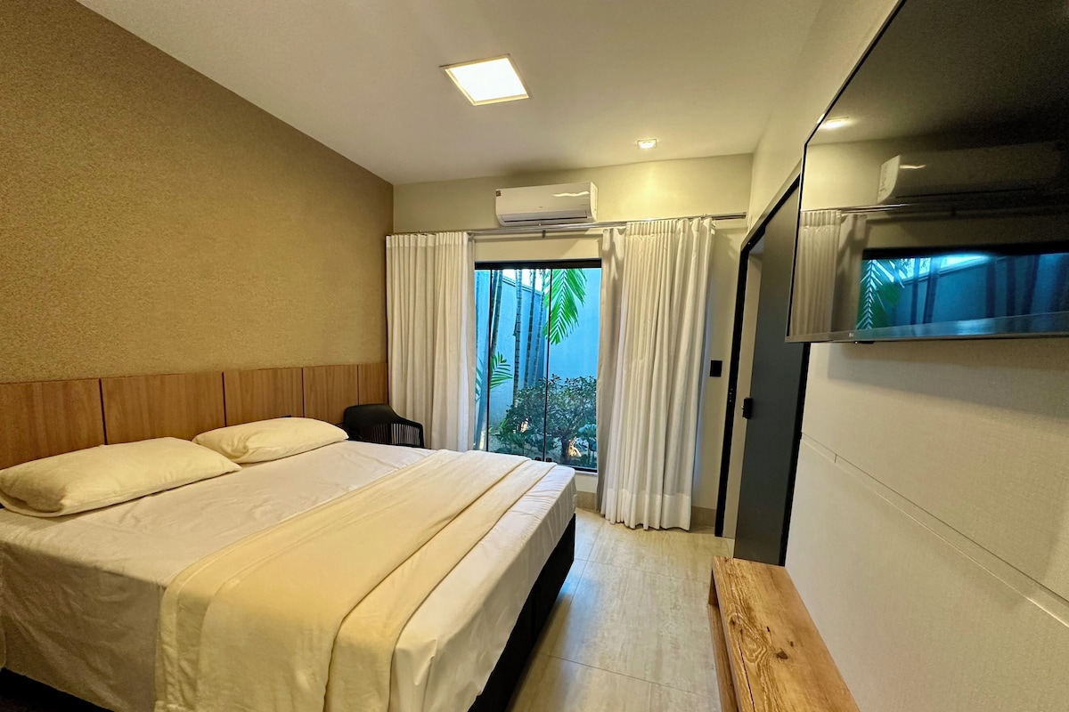 Quarto de hotel com cama, uma mesinha, janela com cortinas e ar condicionado.