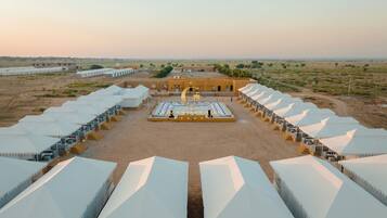 Um resort no deserto com várias barracas brancas dispostas em fileiras, um edifício central com a inscrição 'GRANDINARA RESORT' e uma paisagem plana e aberta ao fundo.