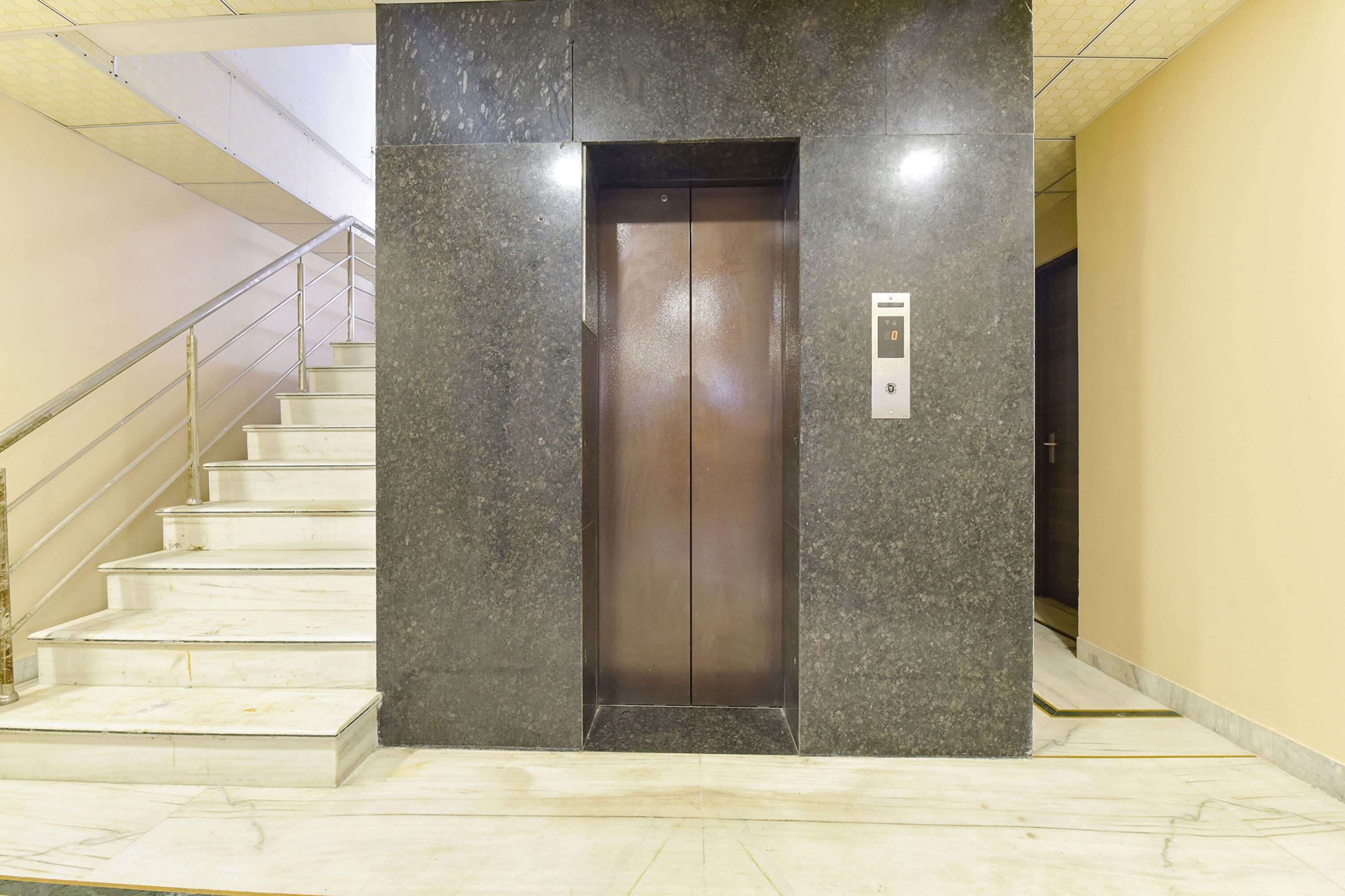 Um elevador em um corredor com piso de mármore e escada de metal.
