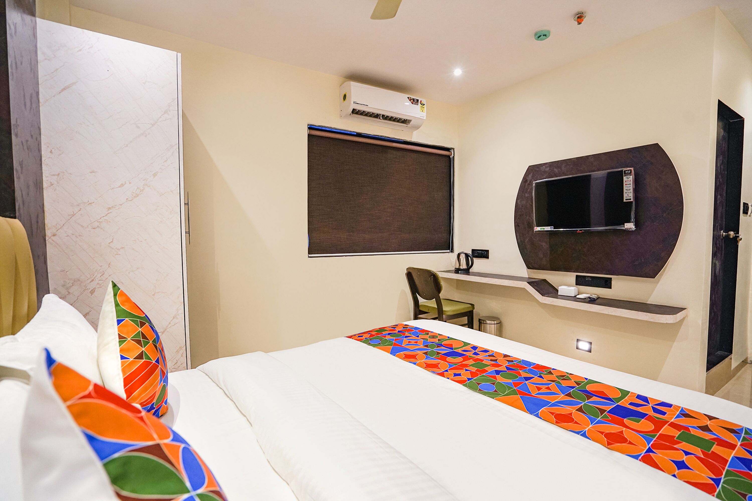 Quarto de hotel com cama, escrivaninha, cadeira, ar condicionado, televisão e porta.