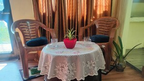 Uma mesa redonda com toalha de renda, duas cadeiras de vime com almofadas, uma panela vermelha com planta e dois copos de vidro sobre a mesa.