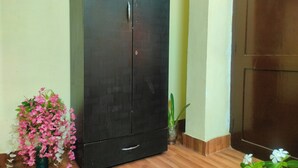 Guarda-roupa de cor escura com puxador, apoiado numa parede amarela com chão de madeira.