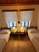 Quarto com duas camas de madeira, cada uma com cabeceira combinando, ladeadas por janelas com cortinas brancas e detalhes em renda. Há duas mesas de cabeceira com abajures e um pequeno objeto decorativo pendurado entre as janelas.