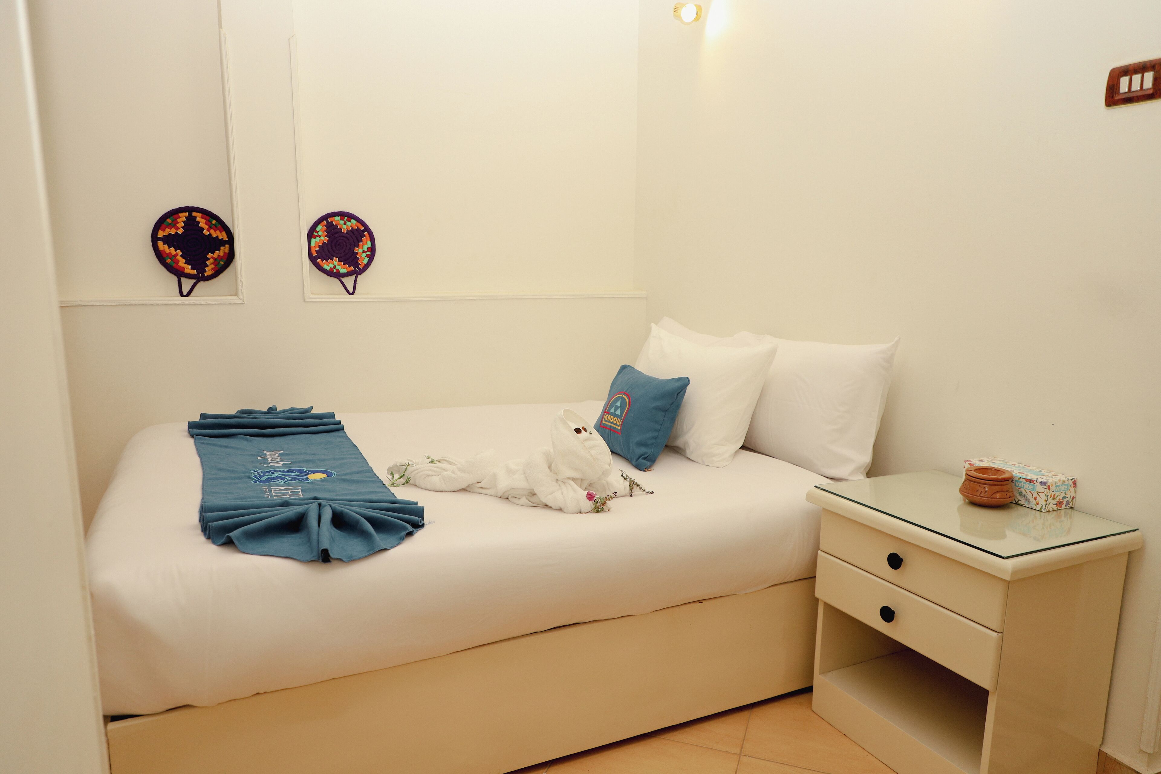Uma cama bem arrumada com manta e almofadas azuis, uma mesinha de cabeceira com tampo de vidro e dois objetos decorativos fixados na parede.