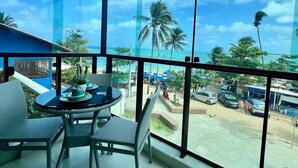 Varanda com mesa e cadeiras, grandes janelas com vista para palmeiras e uma praia.