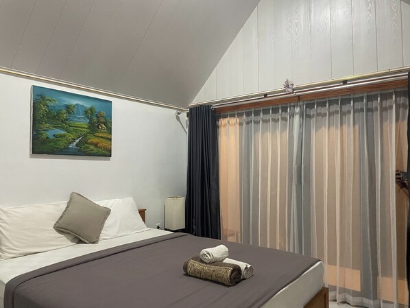 Quarto de hotel com cama, quadro de uma paisagem e janela com cortinas leves.
