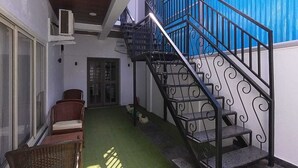 Um corredor exterior estreito com grama sintética verde, escadas com balaústres pretos ornamentados e uma parede azul ao fundo.
