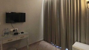 Quarto de hotel com televisão de tela plana na parede, uma pequena mesa com tampo de vidro e um armário branco.