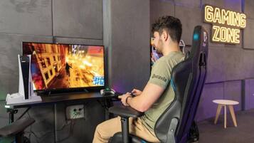 Uma pessoa sentada em uma estação de jogos com monitor exibindo um jogo, uma cadeira gamer e um letreiro neon com a inscrição