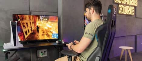 Uma pessoa sentada em uma estação de jogos com monitor exibindo um jogo, uma cadeira gamer e um letreiro neon com a inscrição