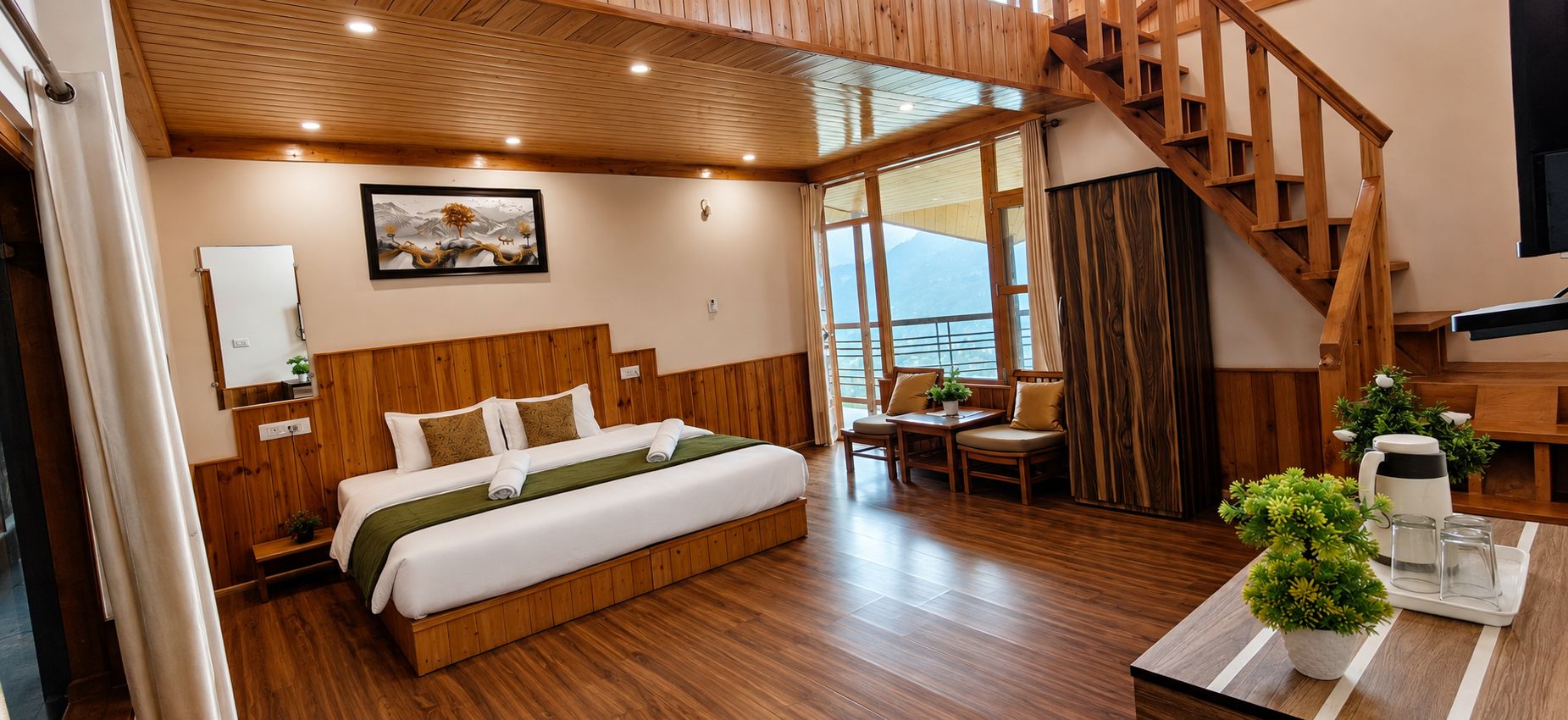 Quarto de hotel com cama de madeira, escrivaninha, cadeira, mesinha, planta em vaso e vista para as montanhas.