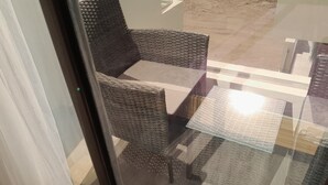 Uma cadeira de vime e uma mesa de vidro num quarto com vista para uma área arenosa.