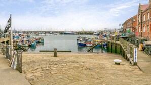 Um porto com barcos atracados, um calçadão de pedras e um edifício com a placa 'Queen Ferry'.