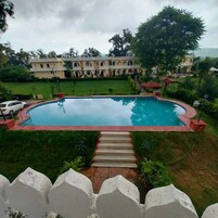 Uma piscina com borda vermelha, rodeada por um gramado e um edifício com várias arcadas ao fundo.