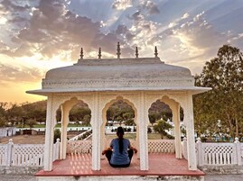 Uma pessoa sentada em um banco sob um pavilhão com teto em cúpula e três torres, cercada por uma cerca branca e árvores.
