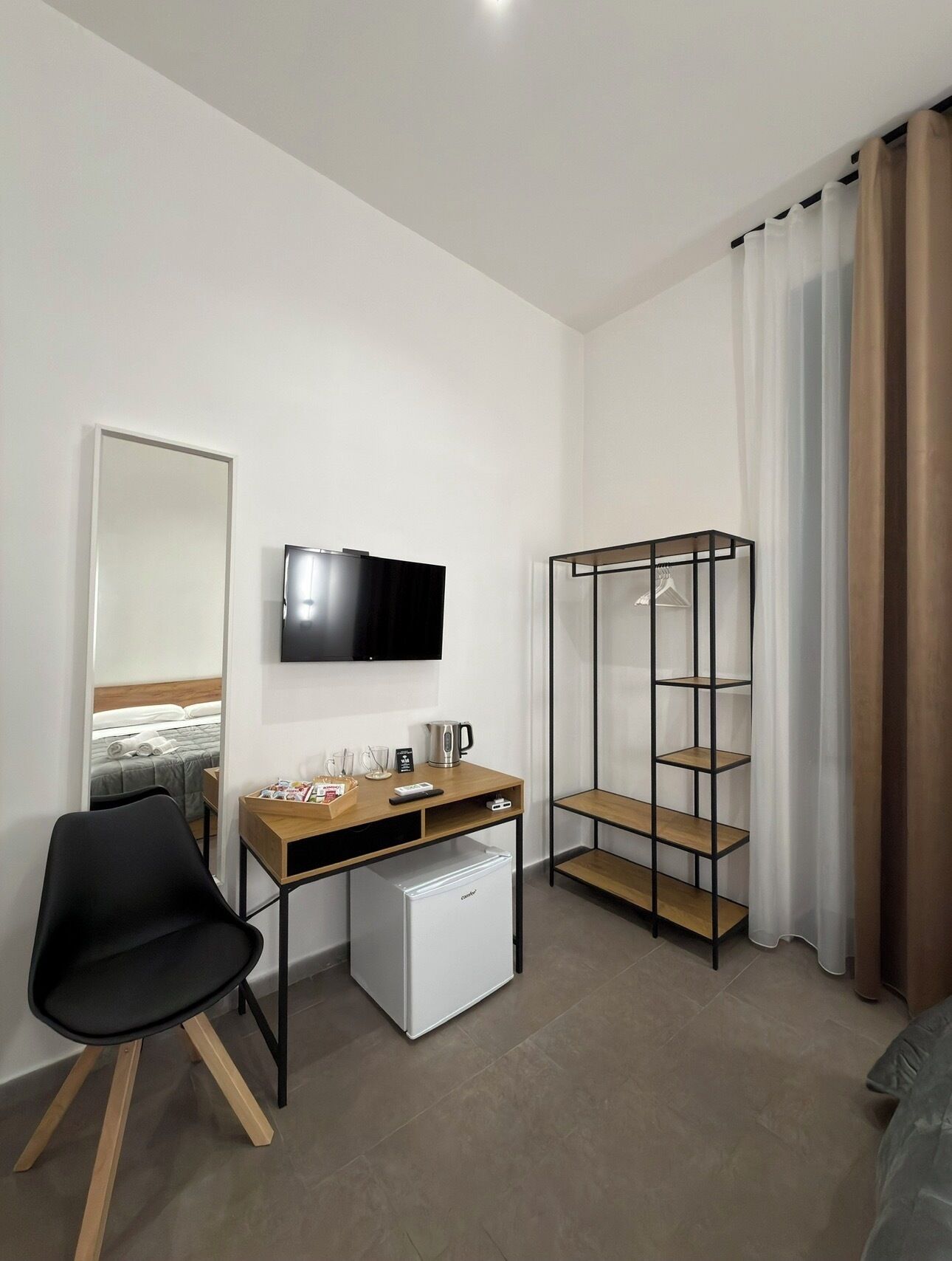 Quarto de hotel moderno com televisão de tela plana, escrivaninha de madeira com mini-geladeira, cadeira preta, armário com prateleiras e cama com cabeceira cinza.