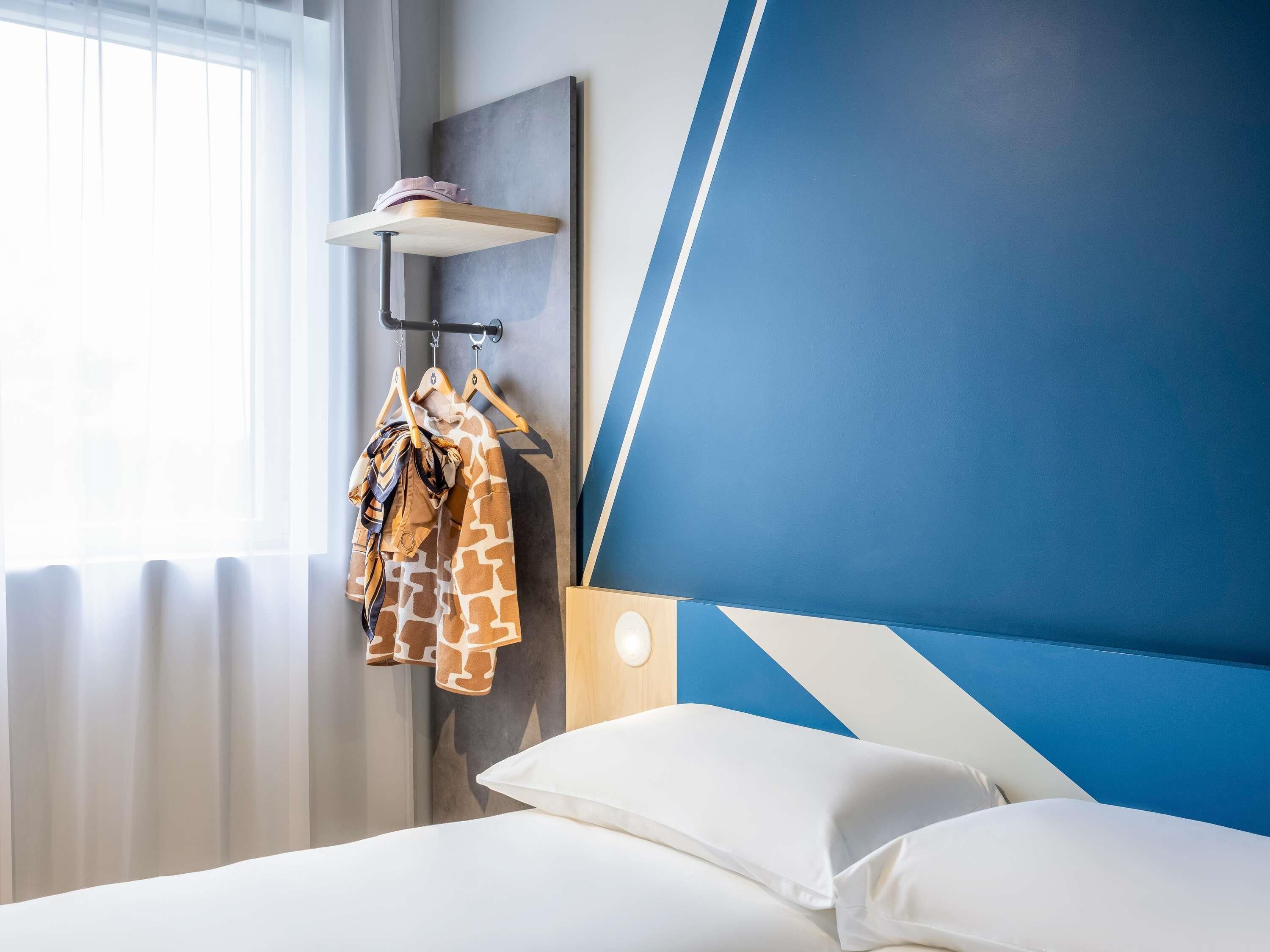 Quarto de hotel com cama, cabeceira, cabideiro fixado na parede com um casaco estampado com girafas e uma janela com cortinas.