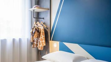 Quarto de hotel com cama, cabeceira, cabideiro fixado na parede com um casaco estampado com girafas e uma janela com cortinas.