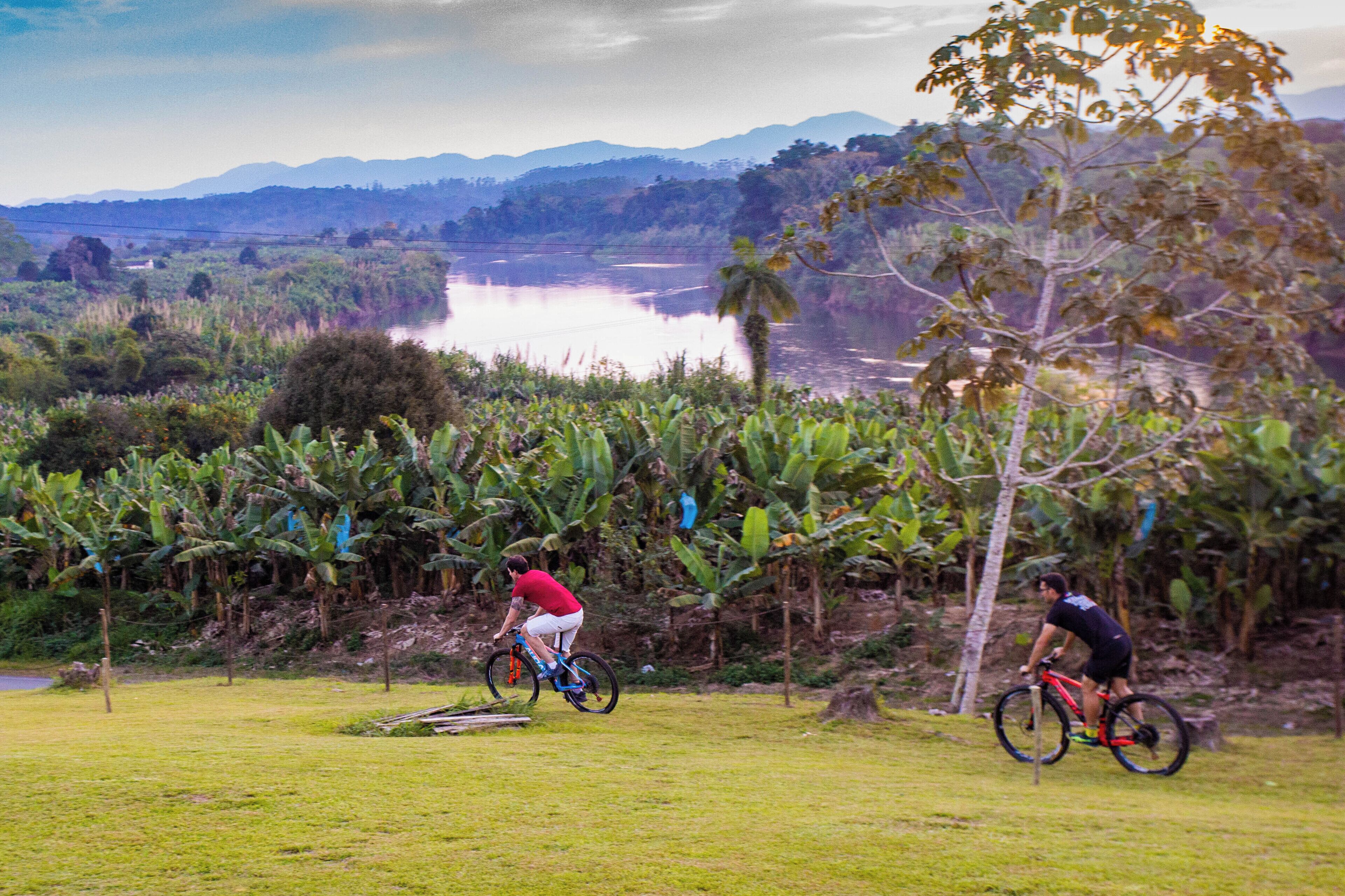 Dois ciclistas em um campo gramado com um rio e montanhas ao fundo.