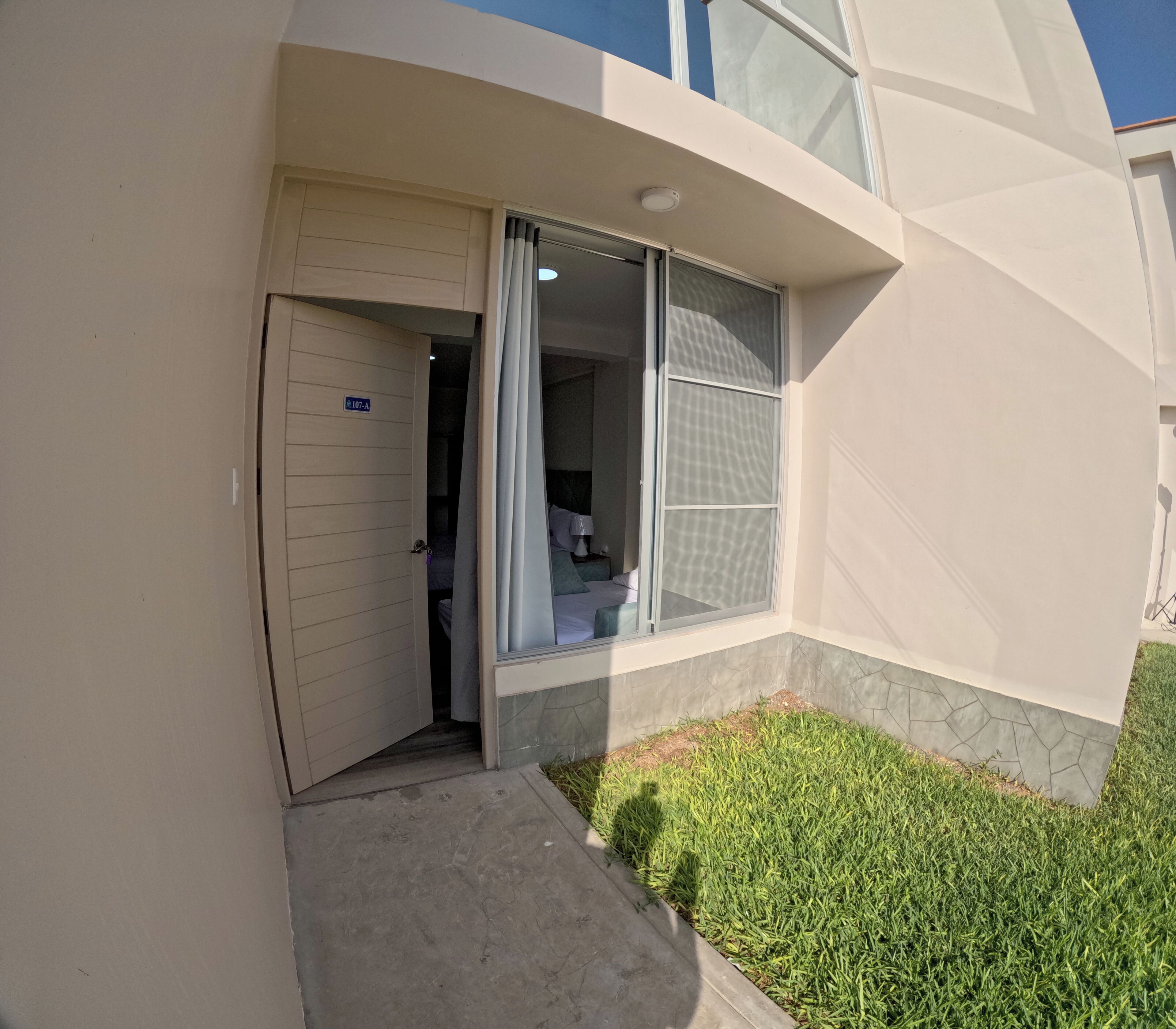 Entrada de um hotel com uma porta identificada como "107-A", uma janela com persiana quadriculada e um pequeno gramado na frente.