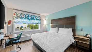 Quarto de hotel com uma cama grande, uma escrivaninha, uma cadeira, uma televisão e uma janela com vista para um edifício e vegetação.