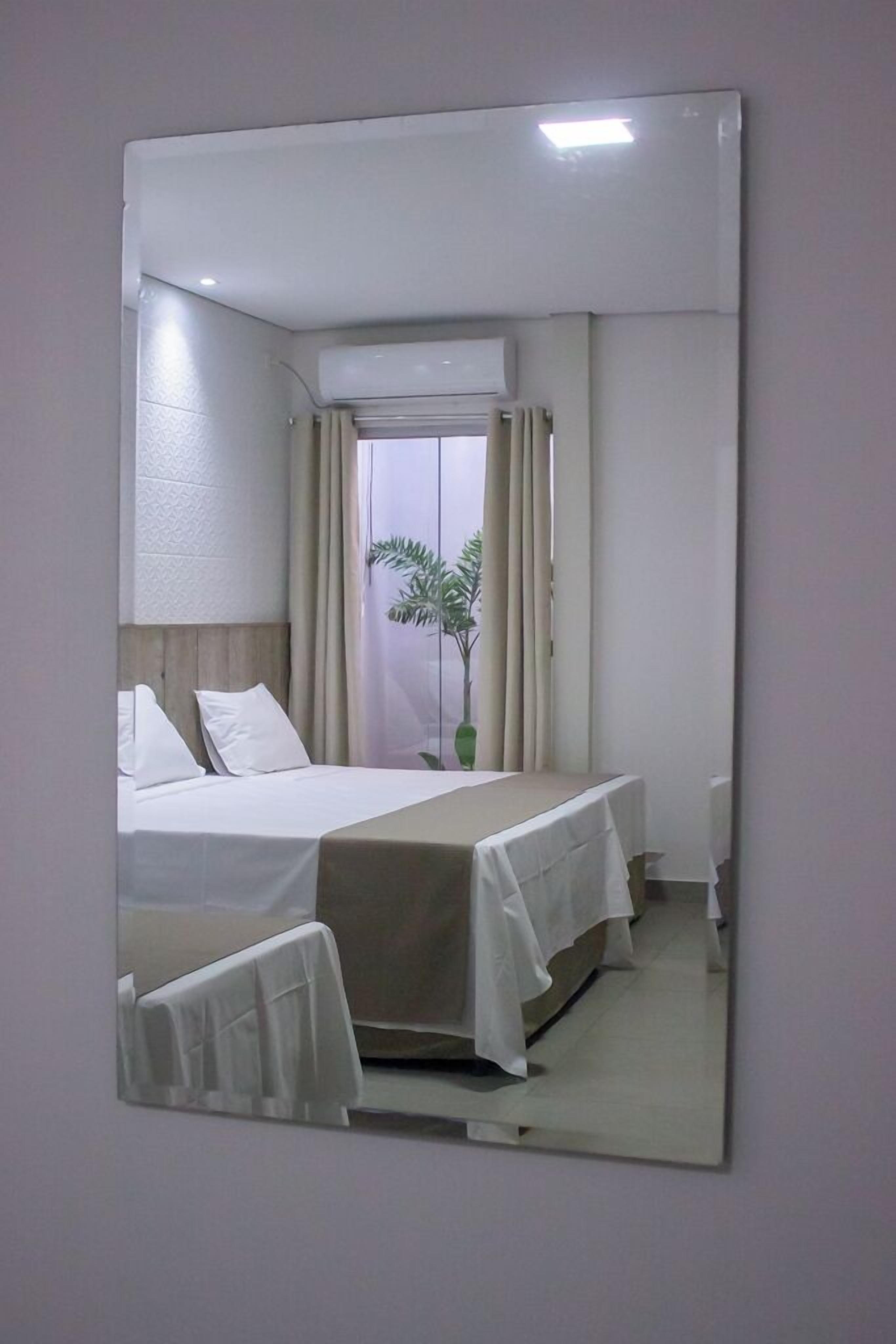 Quarto de hotel com cama, mesa de cabeceira, espelho, janela com cortinas e planta em vaso.