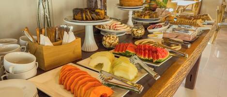 Buffet com diversos pratos e frutas, incluindo melancia e cenouras, dispostos em uma mesa longa.