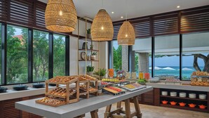 Disposição de buffet com diversos alimentos e vista para o mar através de amplas janelas.