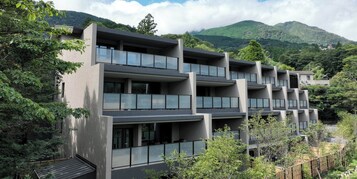 Um edifício moderno de vários andares com varandas, cercado por vegetação exuberante e montanhas ao fundo.