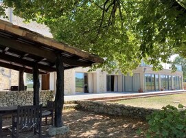 Uma área de estar ao ar livre coberta, com paredes de pedra, pérgola e vista para um edifício moderno com grandes janelas.