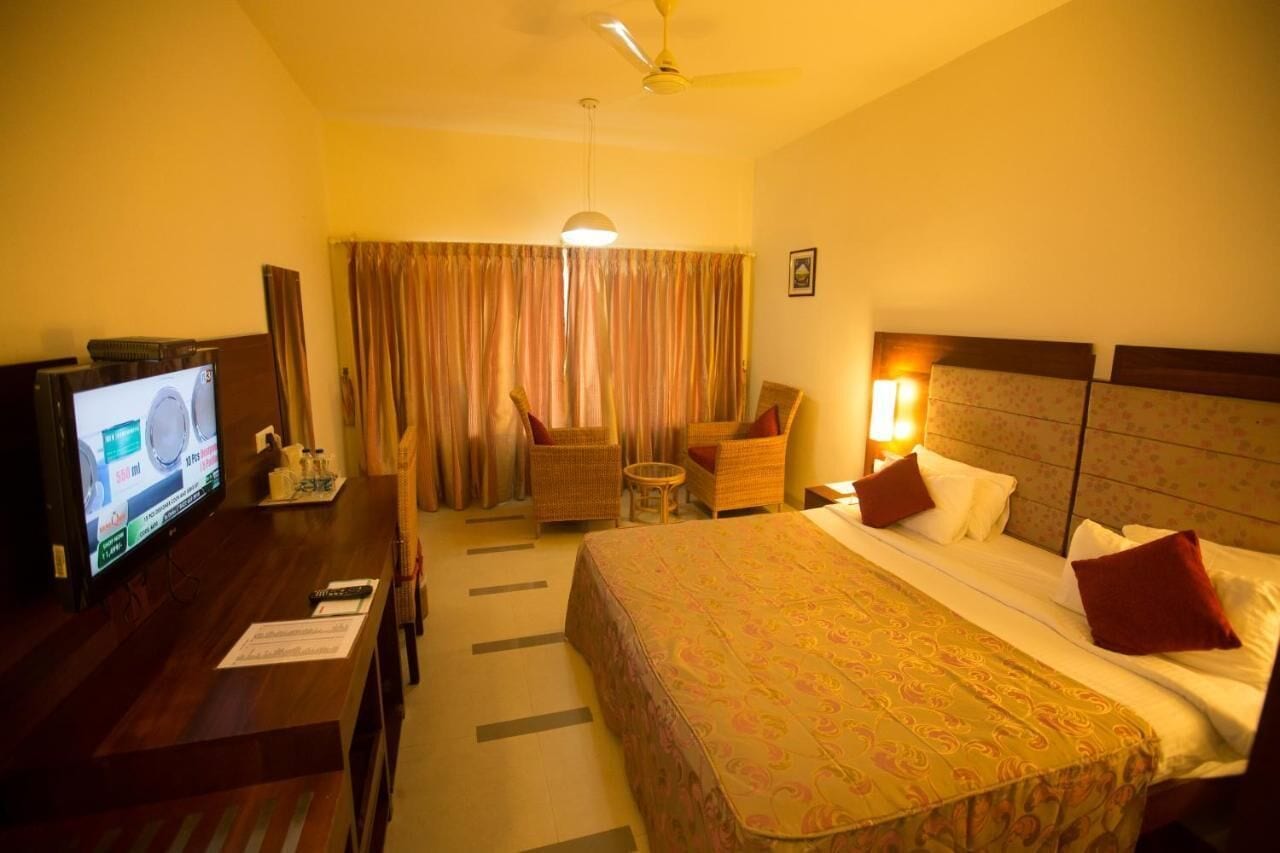 Quarto de hotel com cama, televisão, escrivaninha, cadeira e janela com cortinas.