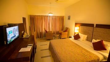 Quarto de hotel com cama, televisão, escrivaninha, cadeira e janela com cortinas.