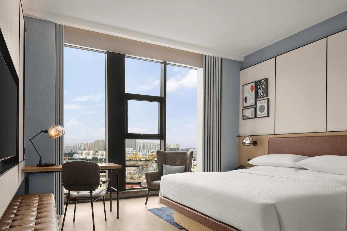 Quarto de hotel moderno com uma cama grande, uma mesa com cadeira e vista da cidade através de janelas amplas, com um horizonte visível.