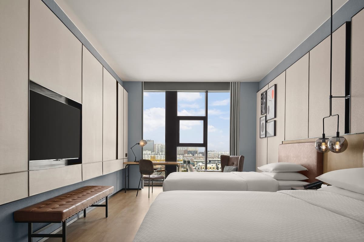 Quarto de hotel moderno com uma cama grande, uma mesa com cadeira, um banco e uma janela ampla com vista para a cidade.