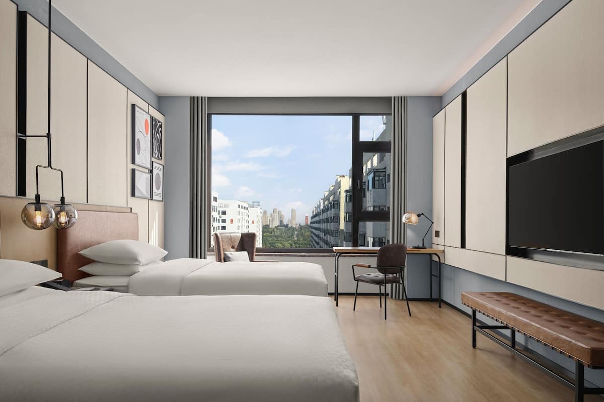 Quarto de hotel moderno com duas camas, uma escrivaninha, uma cadeira, um banco e uma televisão grande.