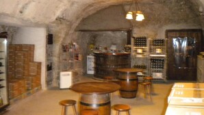 Interior com paredes de pedra, barris de madeira, banquinhos e prateleiras com garrafas.