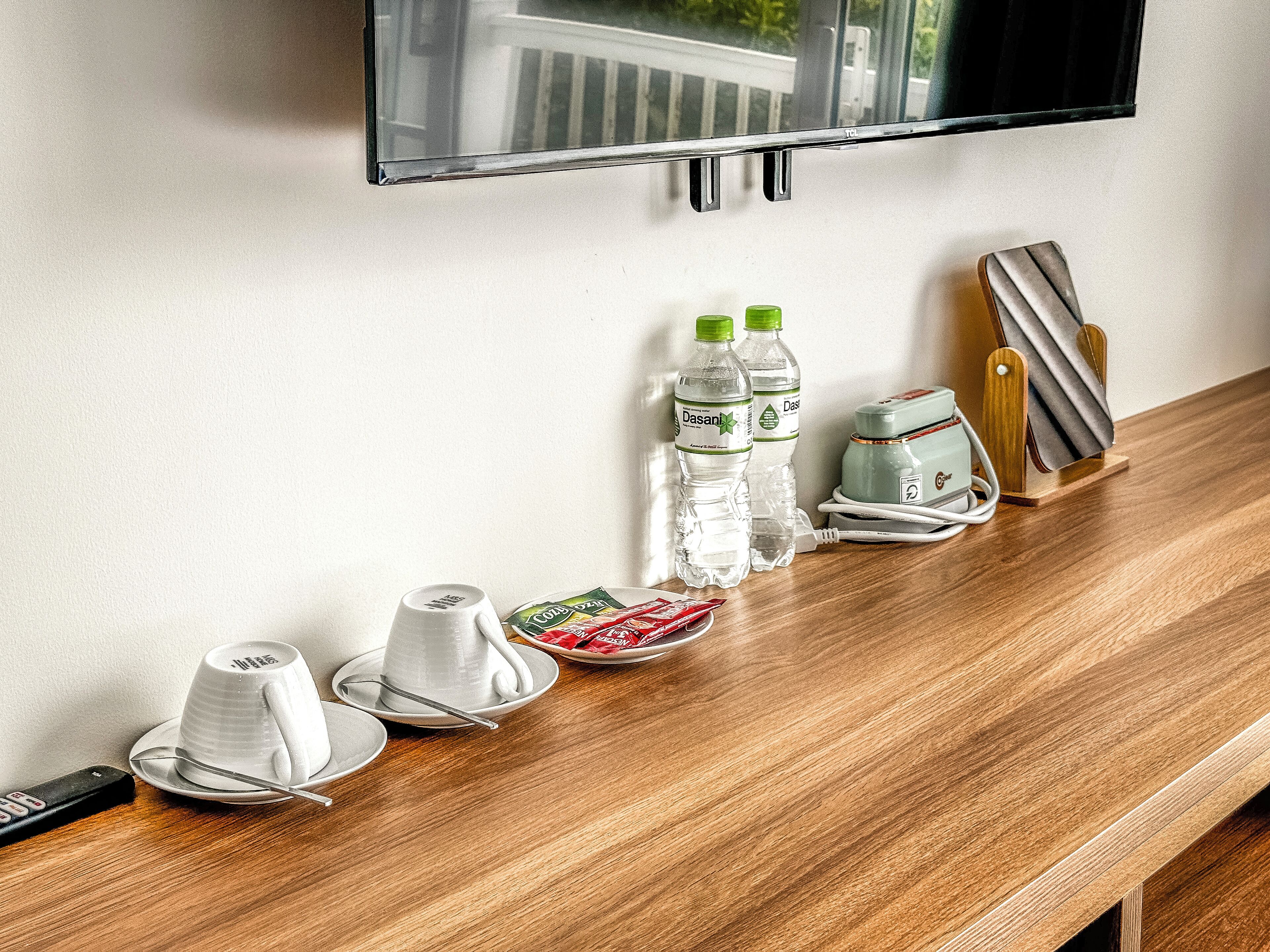 Uma mesa de madeira com um controle remoto, uma xícara branca, um prato com aperitivos, duas garrafas de água Dasani, uma cafeteira verde e um bloco de facas.