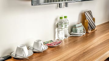 Uma mesa de madeira com um controle remoto, uma xícara branca, um prato com aperitivos, duas garrafas de água Dasani, uma cafeteira verde e um bloco de facas.