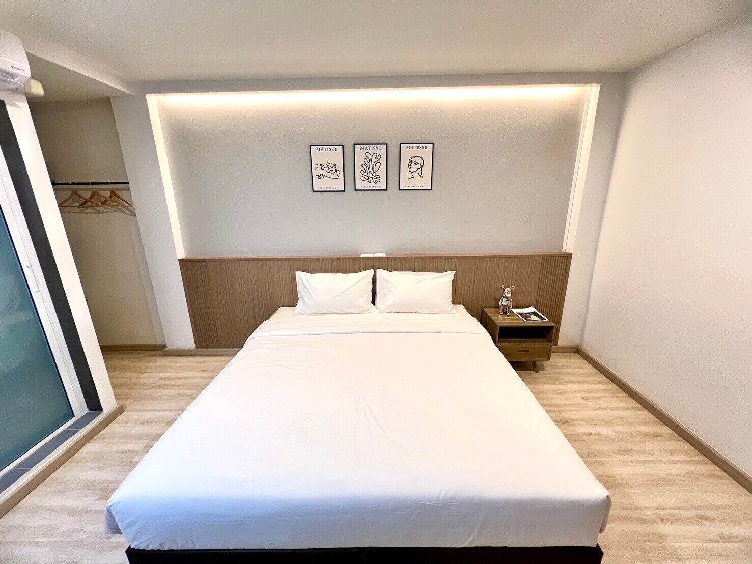 Quarto de hotel moderno com uma cama grande, cabeceira de madeira e uma mesa de cabeceira com abajur. Três obras de arte emolduradas na parede acima da cama.