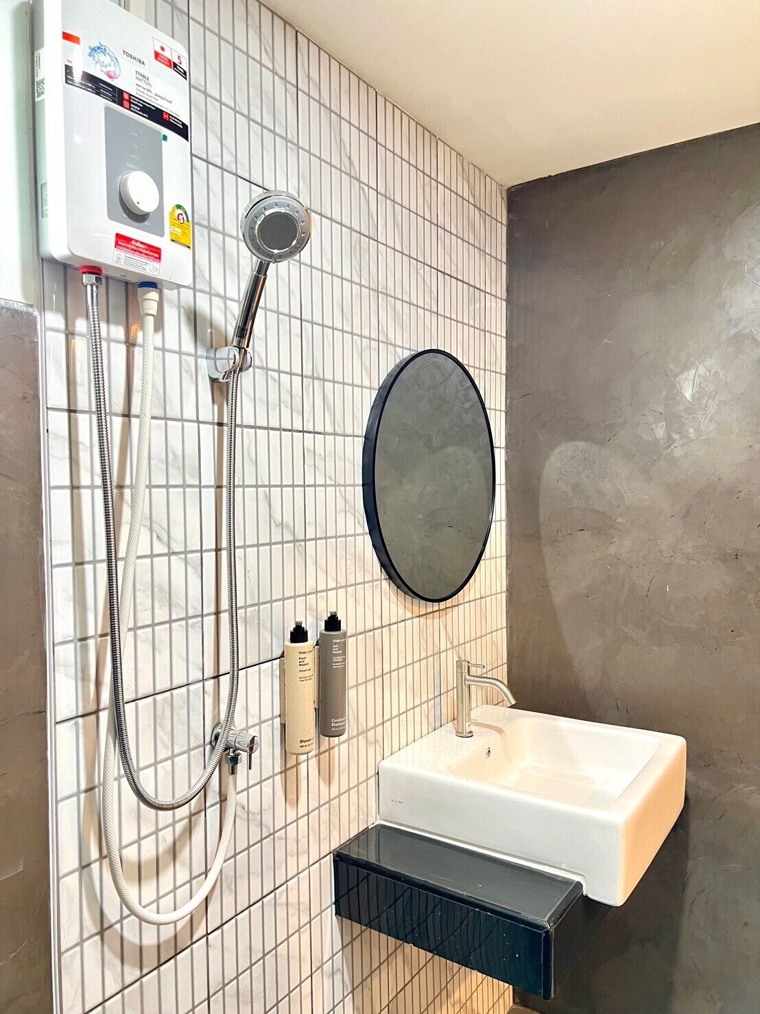 Casa de banho com parede revestida a azulejos brancos, lavatório preto, espelho e chuveiro manual.