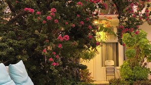 Um jardim com um canteiro de flores azuis claras, uma cadeira branca e um edifício com janela e porta ao fundo.