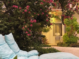 Um jardim com um canteiro de flores azuis claras, uma cadeira branca e um edifício com janela e porta ao fundo.