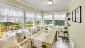 Uma área de refeições com uma mesa de madeira comprida, bancos acolchoados e grandes janelas com cortinas leves.