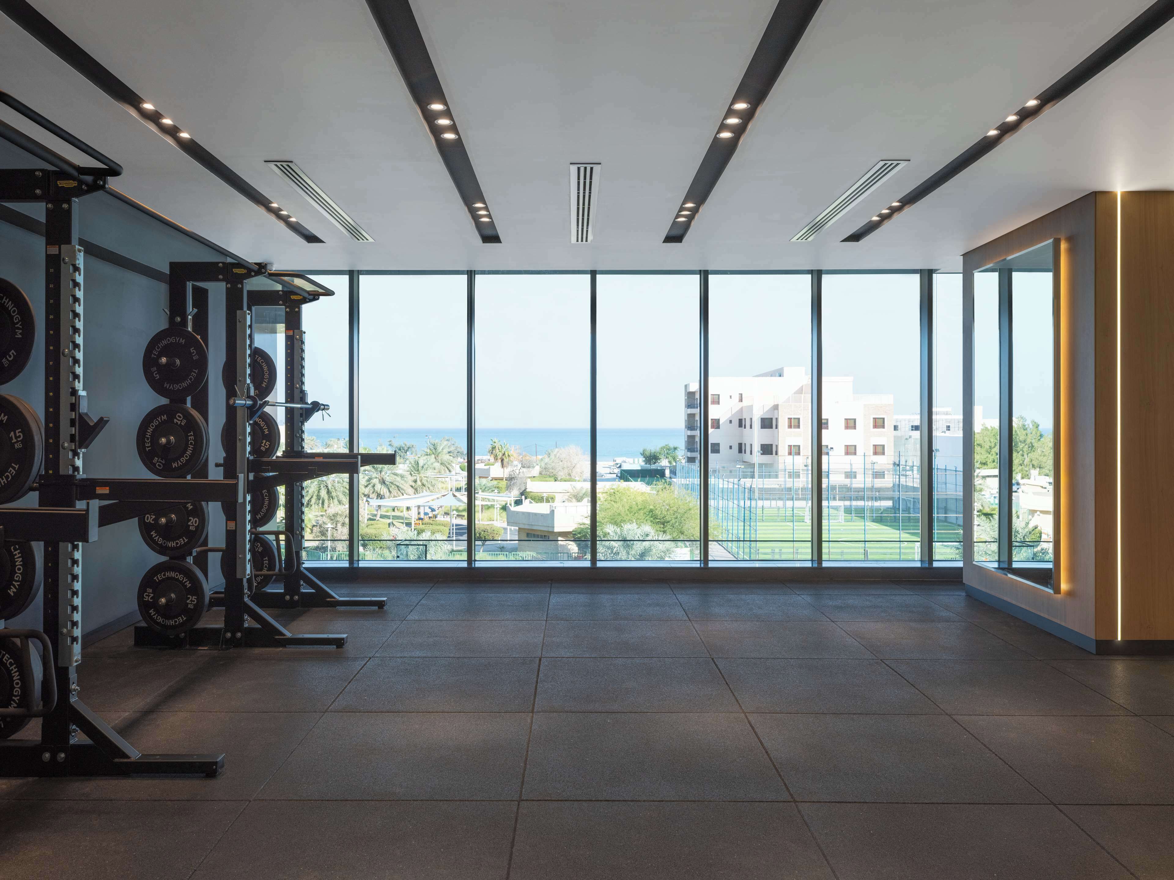 Uma academia com equipamentos de musculação e amplas janelas que oferecem vista para o mar e edifícios.