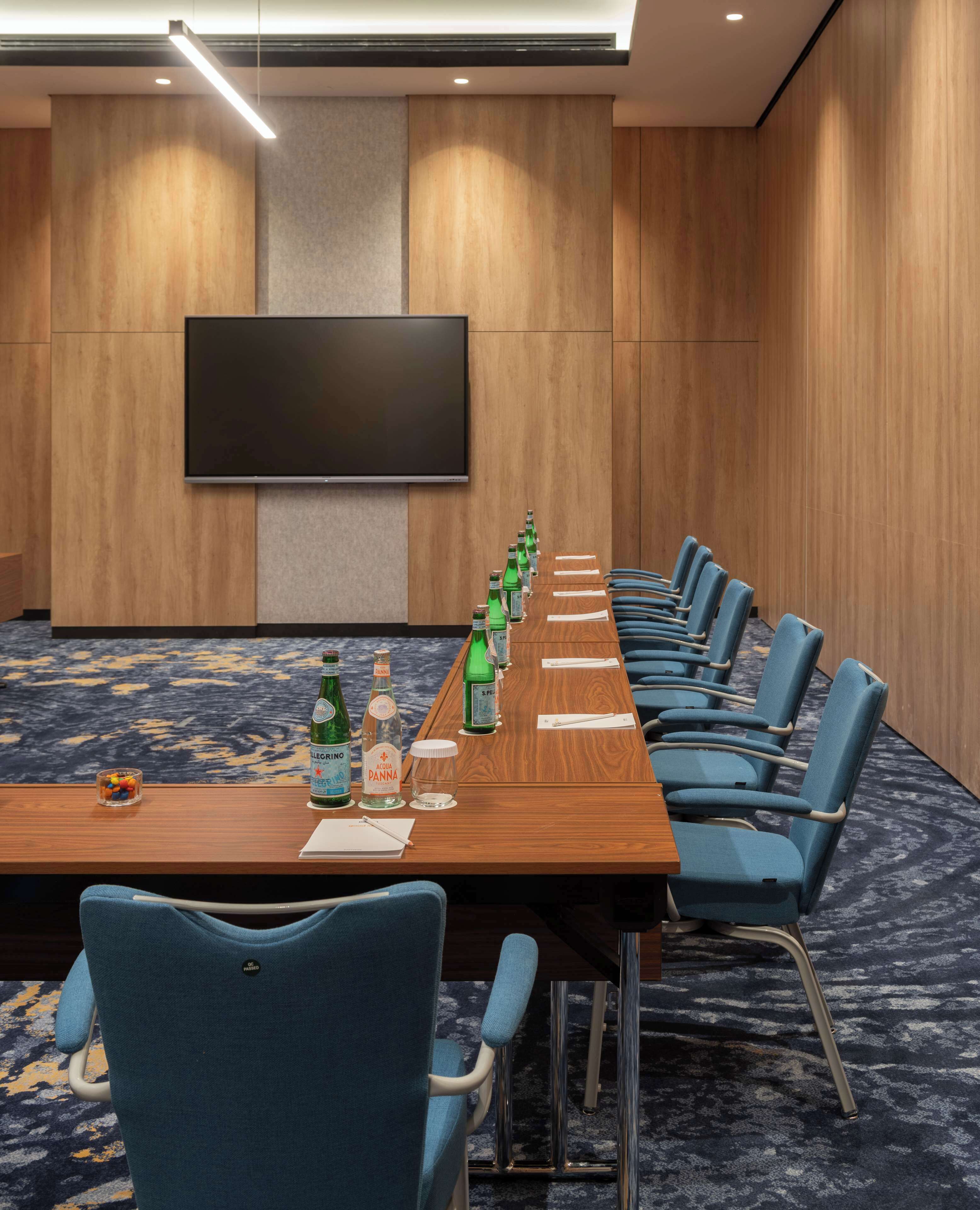 Uma sala de conferências com uma mesa comprida, cadeiras azuis e uma televisão de tela plana na parede.