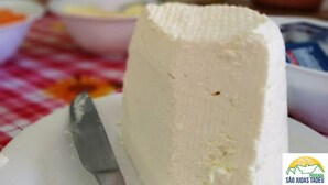 Close-up de um queijo branco num prato, com uma faca, rodeado por diversos acompanhamentos e pratos numa mesa.