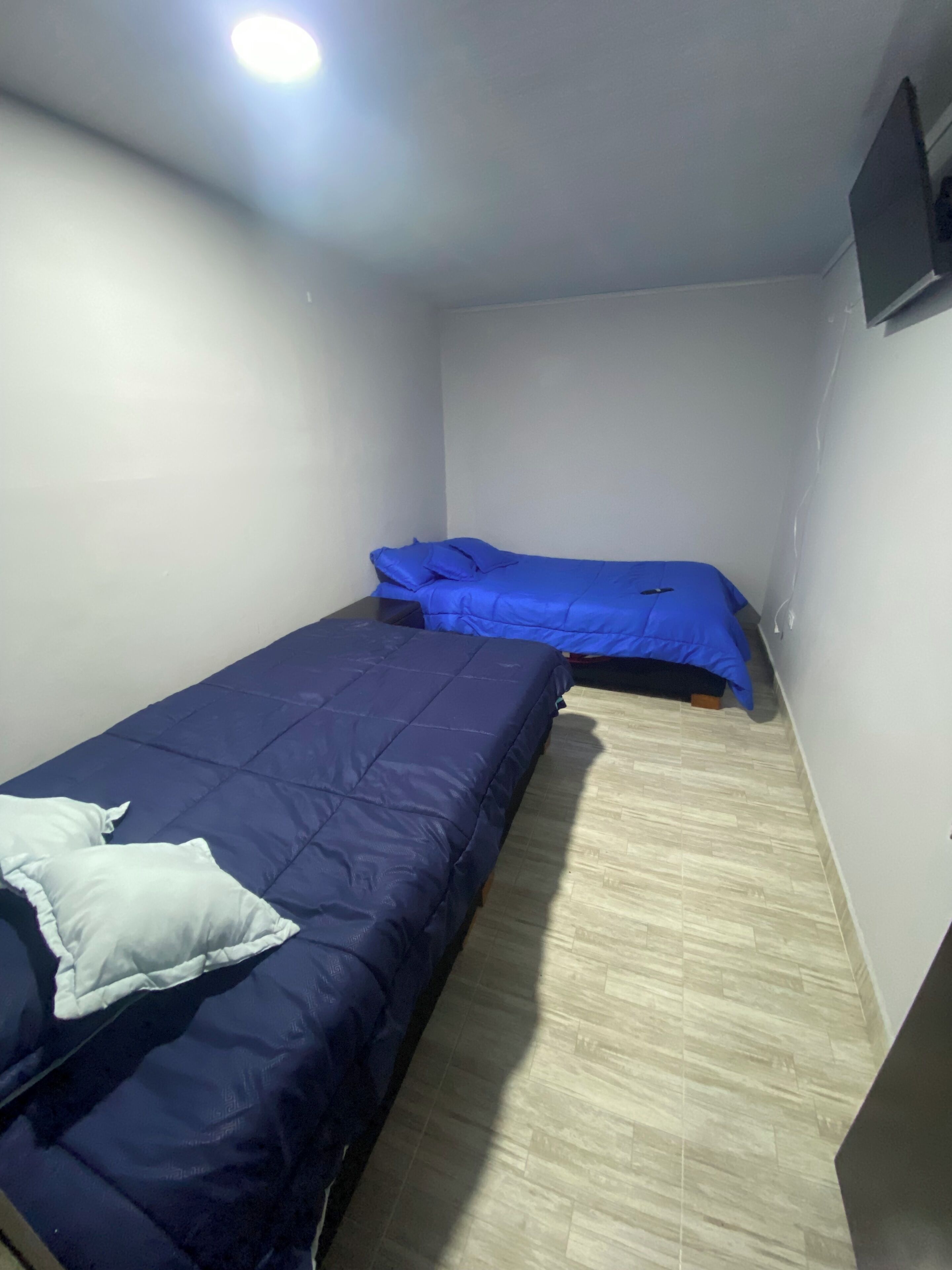 Quarto pequeno e único, com duas camas, uma com cobertura azul e a outra com cobertura escura.